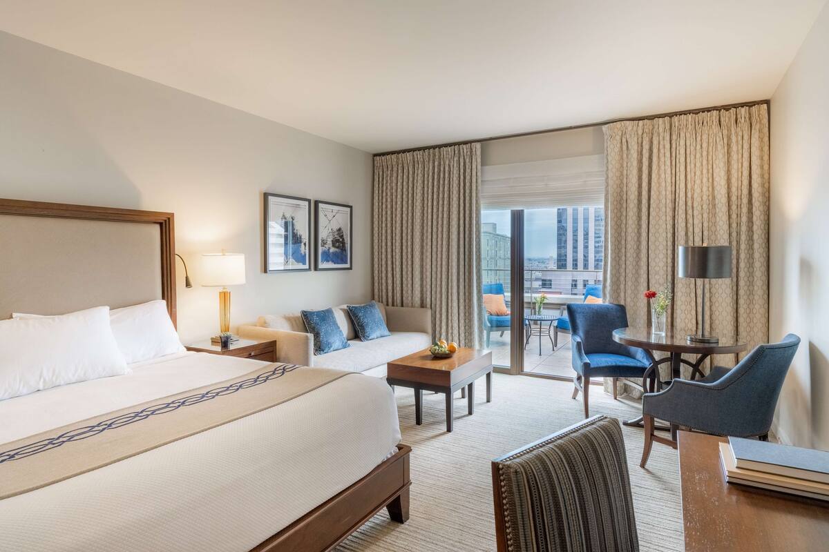 Quarto de hotel com uma cama grande, um sofá, uma escrivaninha e uma cadeira. Há uma janela com cortinas e vista para edifícios.