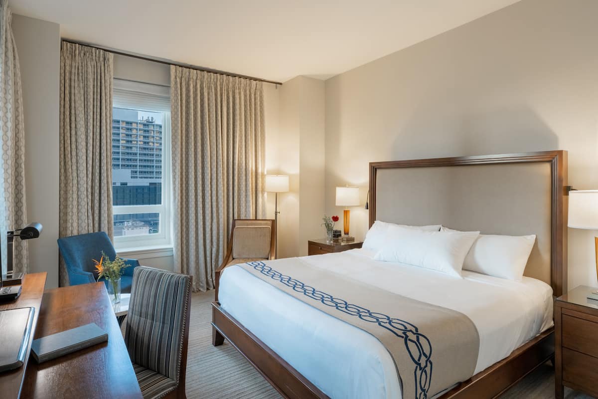Quarto de hotel com uma cama grande, uma escrivaninha, uma cadeira e uma janela com cortinas.