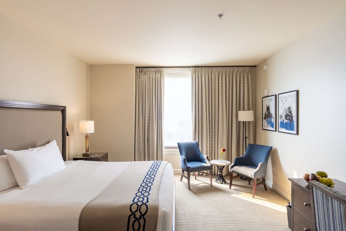Quarto de hotel com uma cama grande, duas cadeiras, uma mesinha, um abajur e uma cômoda com frutas.