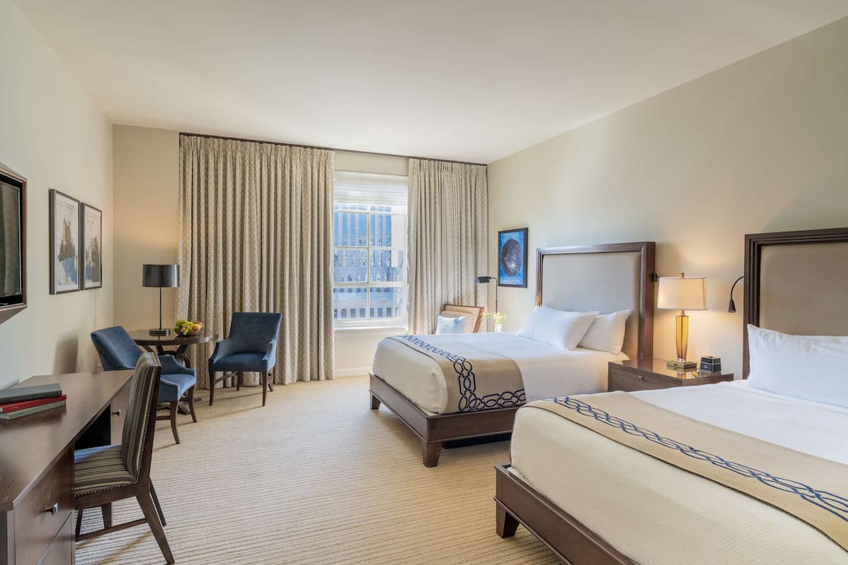 Quarto de hotel com duas camas, uma escrivaninha, uma cadeira, um abajur e uma janela com cortinas.