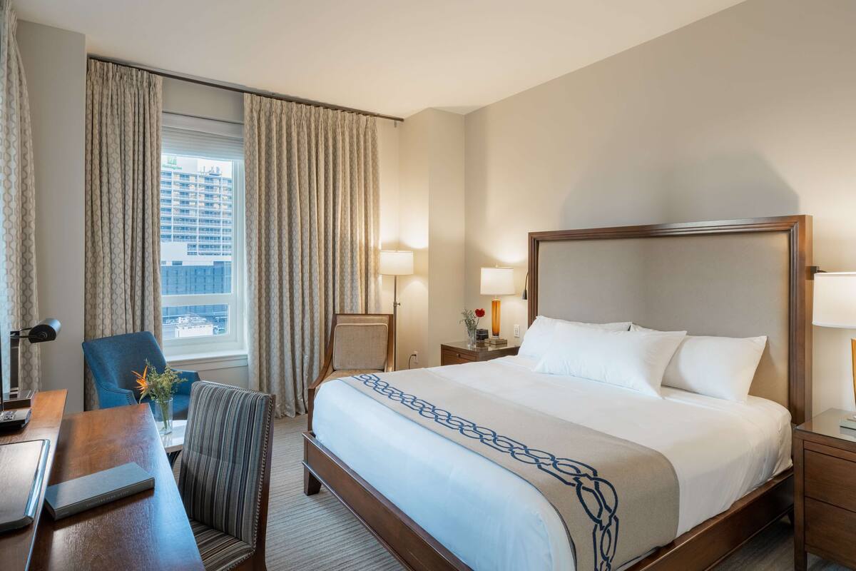 Quarto de hotel com uma cama grande, uma mesa com cadeira, uma luminária e vista para a cidade através da janela.