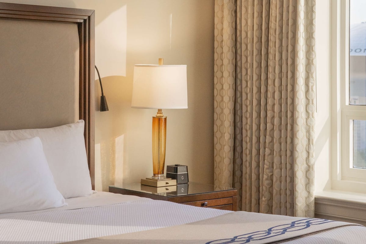 Quarto de hotel com cama, mesa de cabeceira com abajur e janela com cortinas.