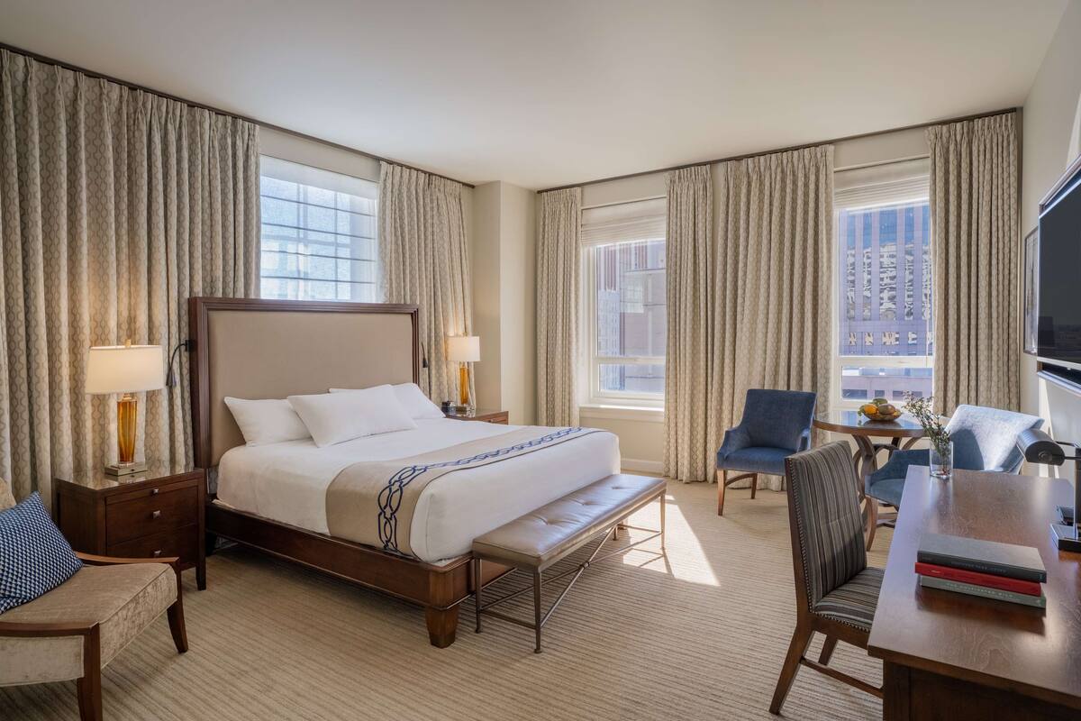 Quarto de hotel com uma cama grande, duas mesas de cabeceira com luminárias, uma escrivaninha com cadeira, uma pequena mesa com um vaso e uma janela grande com cortinas.