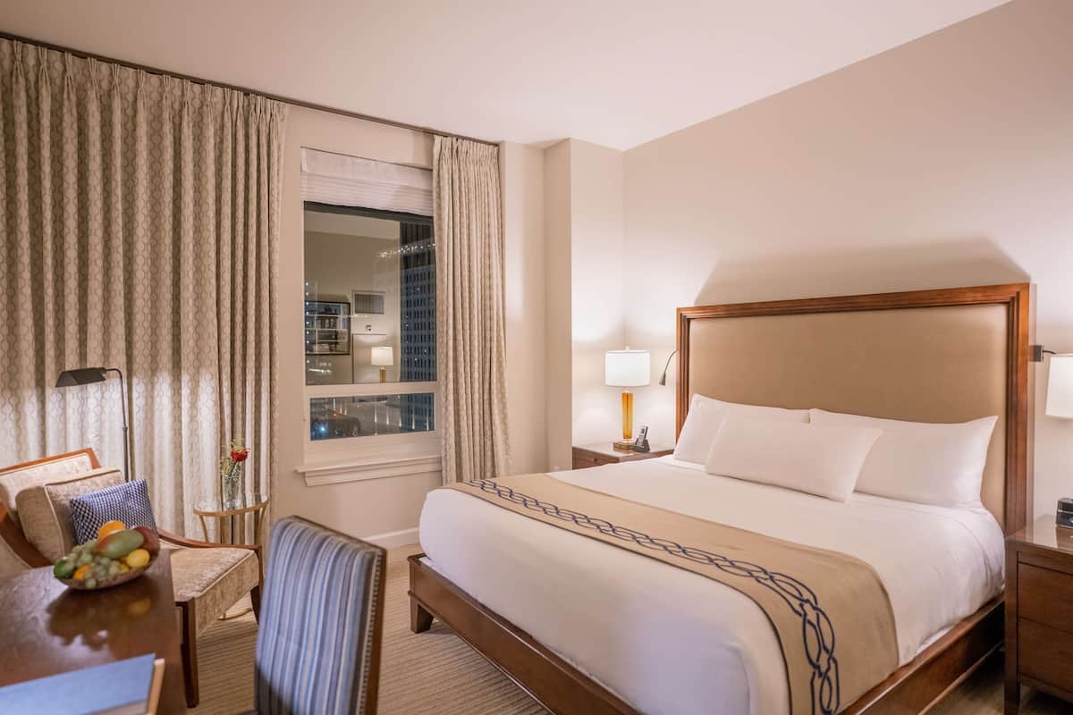 Quarto de hotel com uma cama grande, mesa de cabeceira com abajur, uma cadeira, uma pequena mesa com uma tigela de frutas e uma janela com cortinas.
