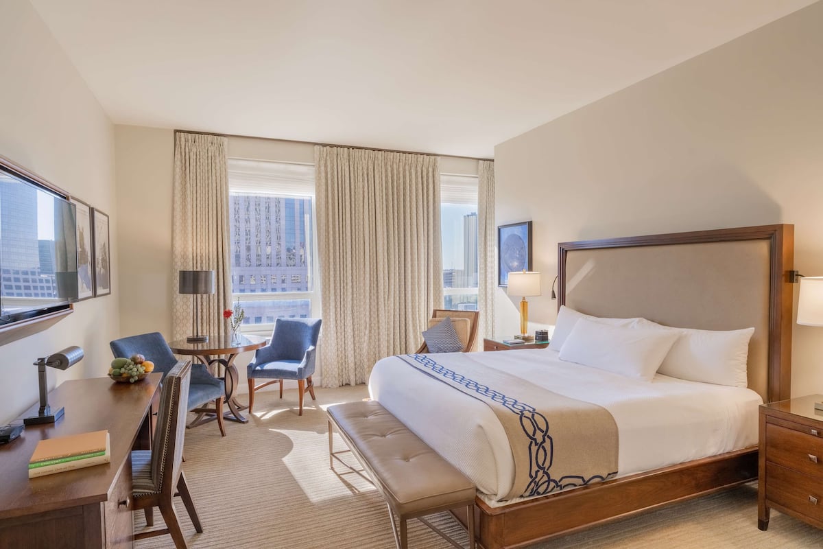 Quarto de hotel com uma cama grande, uma escrivaninha com cadeira, uma mesinha e um banco.