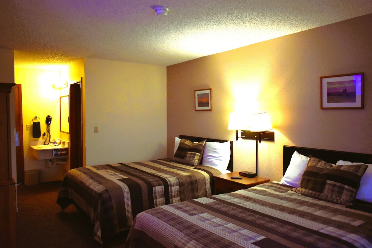 Quarto de hotel com duas camas, uma mesa de cabeceira com abajur, uma pequena escrivaninha com telefone e um quadro na parede.