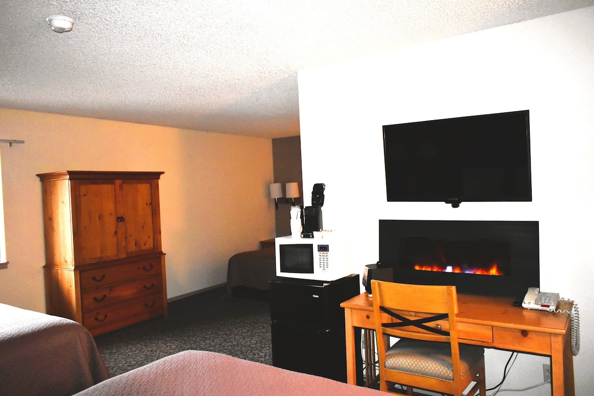 Quarto de hotel compacto com cama, armário de madeira, escrivaninha com cadeira, micro-ondas, televis ão de tela plana e lareira.