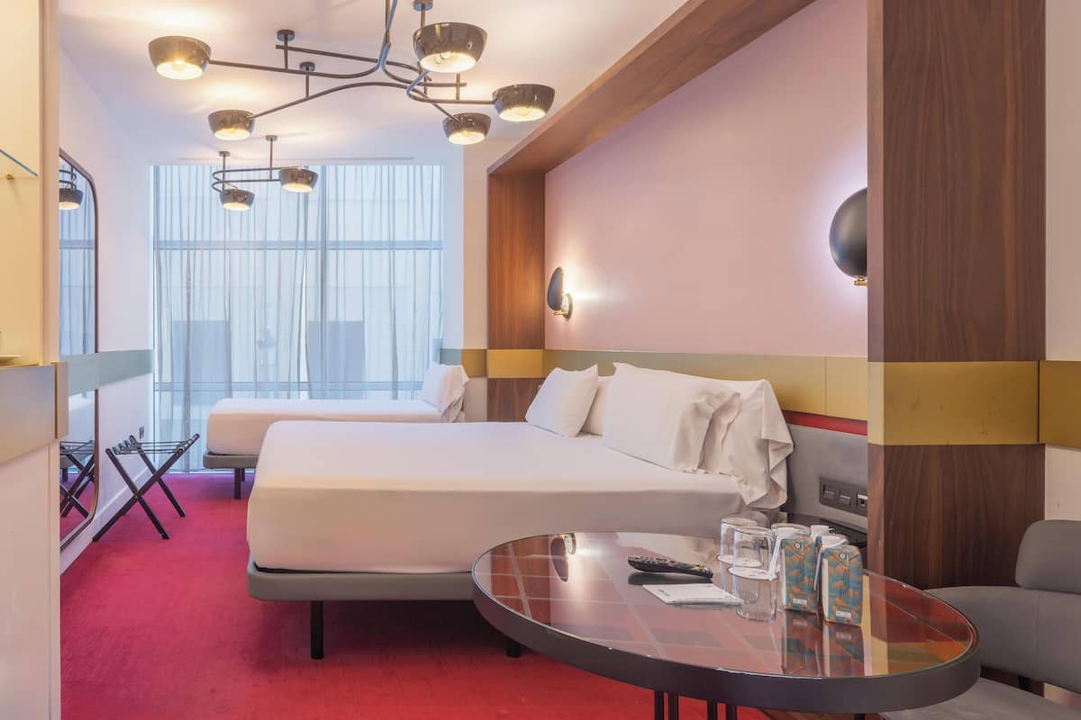 Quarto de hotel com cama, mesa redonda com controle remoto, copos e menu, janela com cortinas e um candelabro.