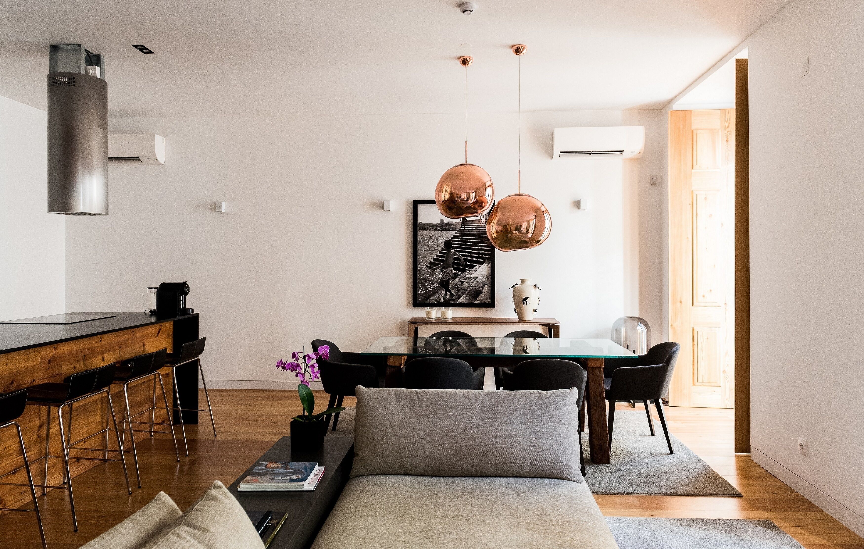 Cozinha moderna com bancada de madeira e banquetas, área de jantar com mesa e cadeiras de vidro, e uma janela grande que proporciona iluminação natural.