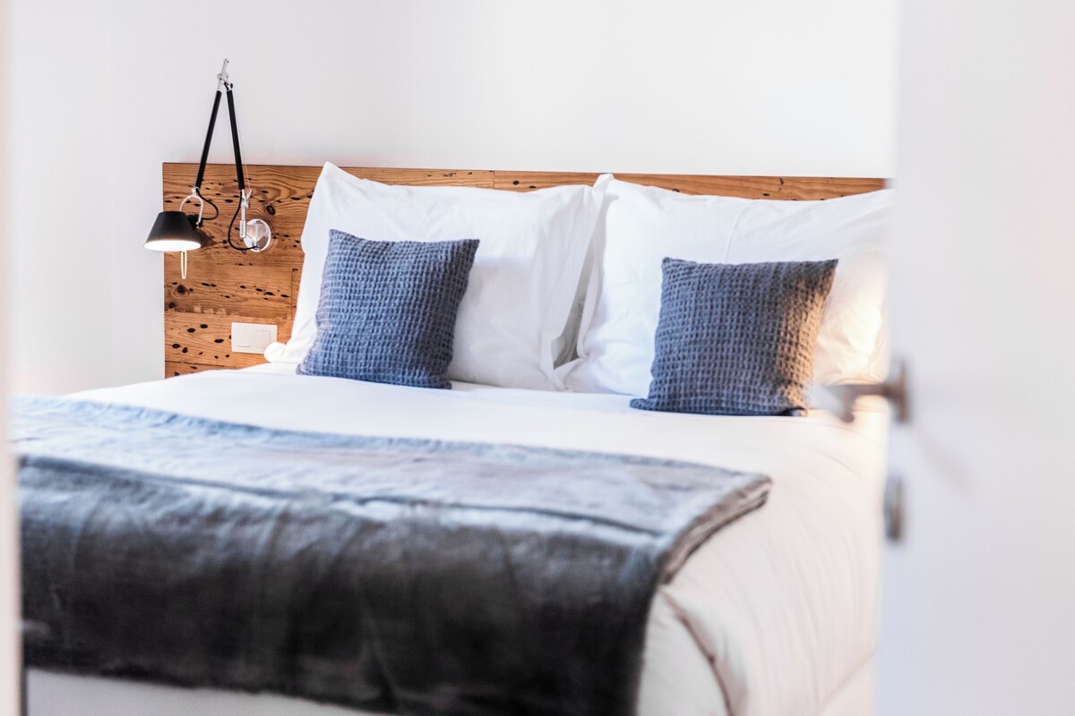 Uma cama bem arrumada com um cobertor branco e azul, duas almofadas e um abajur ao lado.