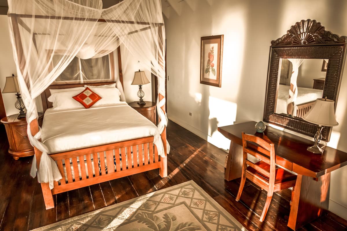 Uma cama com dossel e cortina, uma escrivaninha de madeira com espelho e uma mesa de cabeceira com abajur.