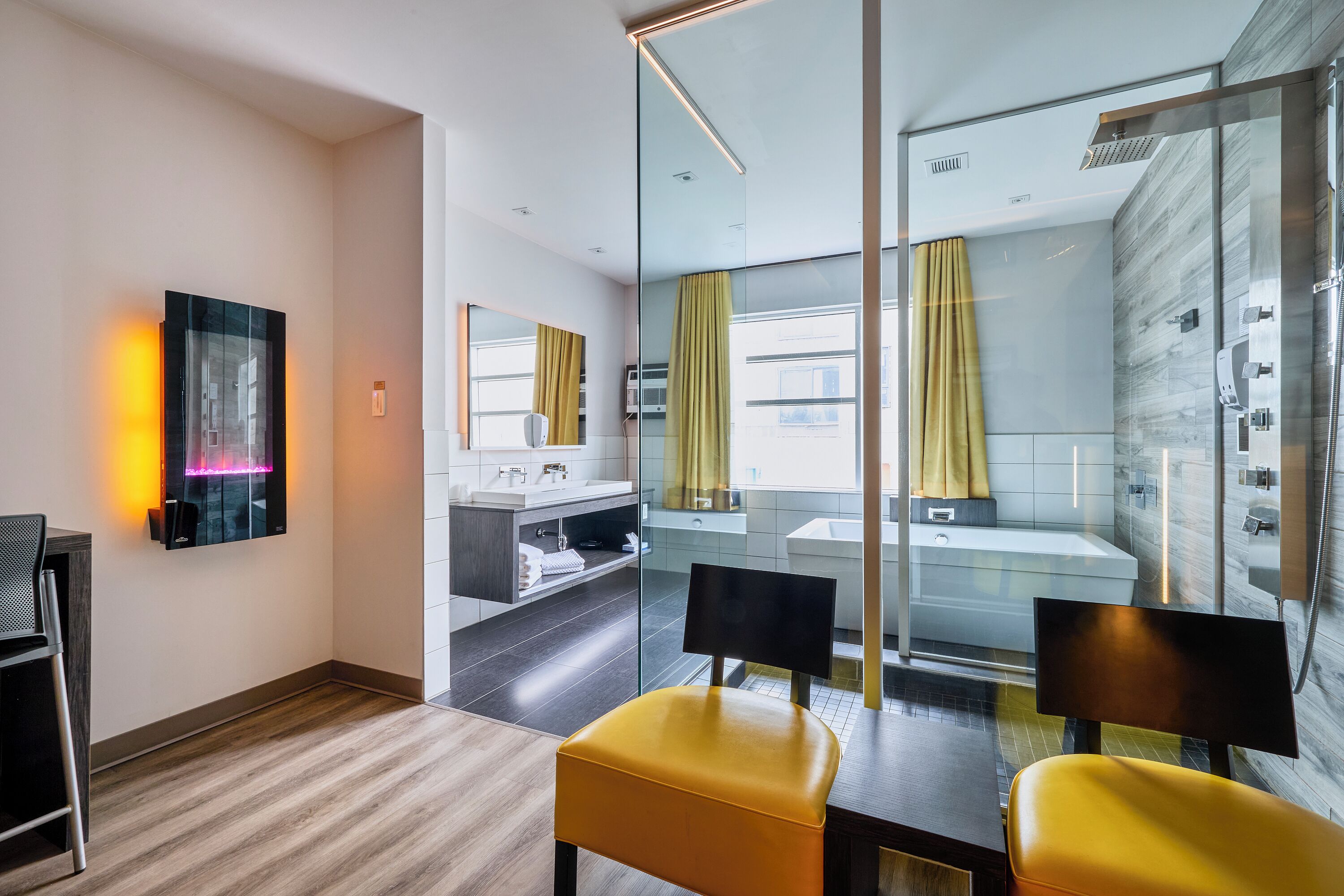 Quarto de hotel moderno com divisória de vidro, um espelho grande, uma cama, mesa de cabeceira, uma cadeira e uma escrivaninha com cadeira.