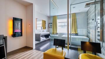 Quarto de hotel moderno com divisória de vidro, um espelho grande, uma cama, mesa de cabeceira, uma cadeira e uma escrivaninha com cadeira.