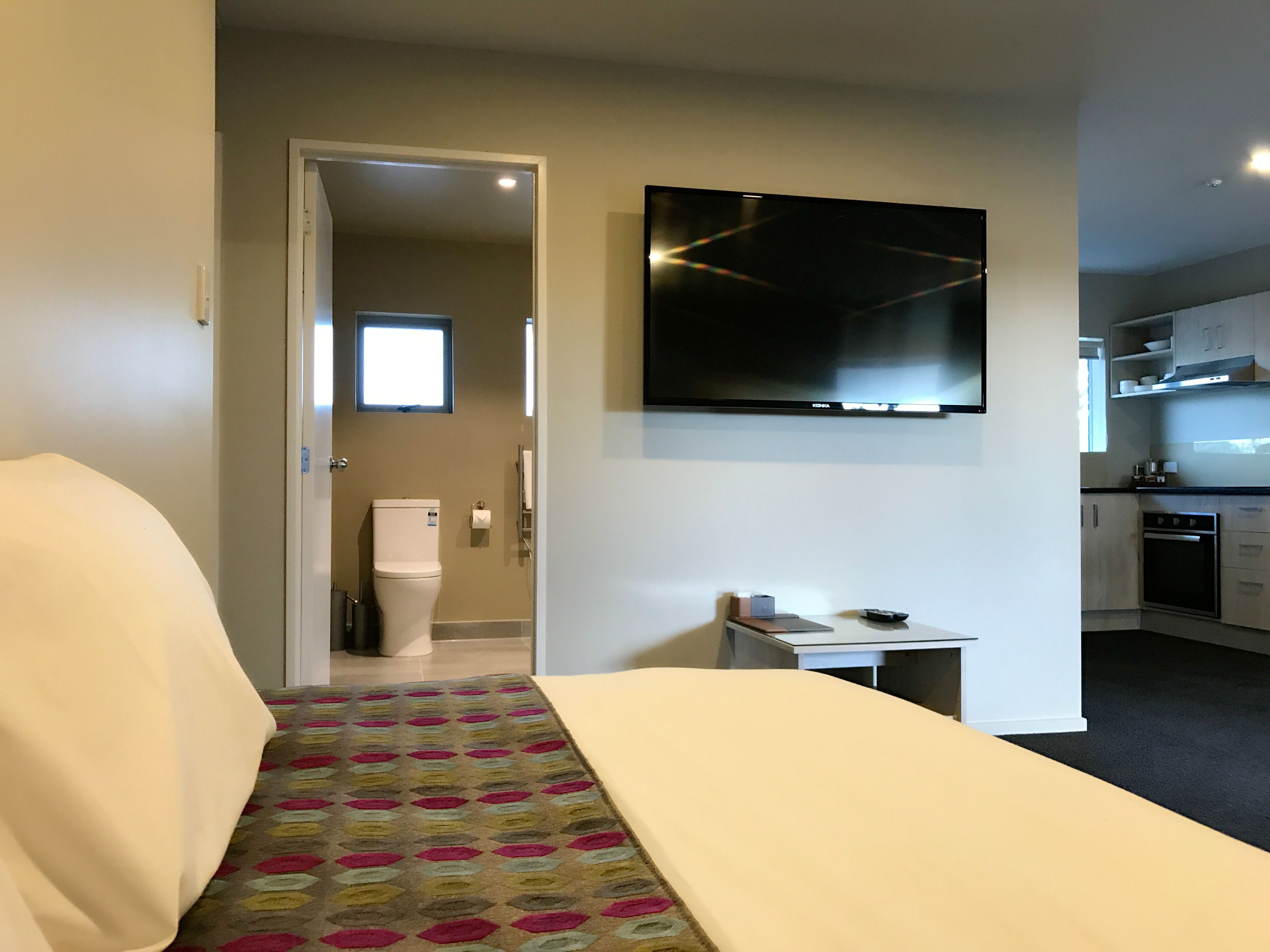 Quarto de hotel com cama, televisão presa na parede, uma mesa pequena e vista para o banheiro e a cozinha.