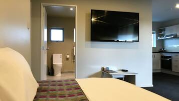 Quarto de hotel com cama, televisão presa na parede, uma mesa pequena e vista para o banheiro e a cozinha.