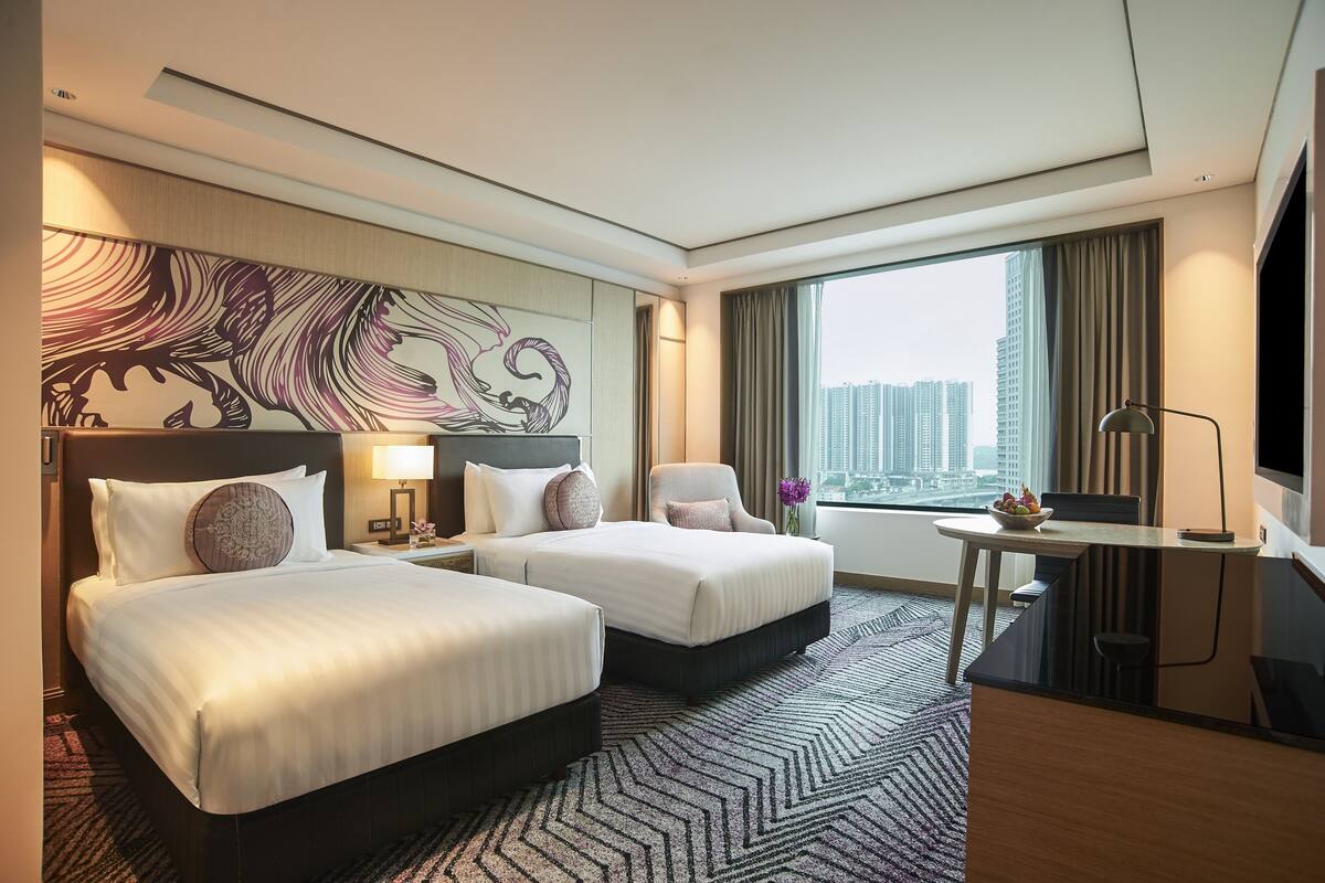 Quarto de hotel com duas camas, uma escrivaninha, televisão e uma janela grande com vista para a cidade.