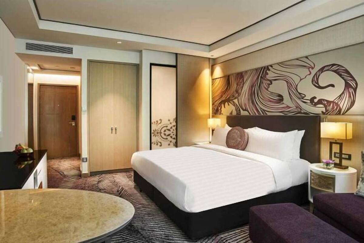 Quarto de hotel com uma cama grande, uma mesa de jantar redonda, uma mesinha lateral e uma parede com uma pintura decorativa.