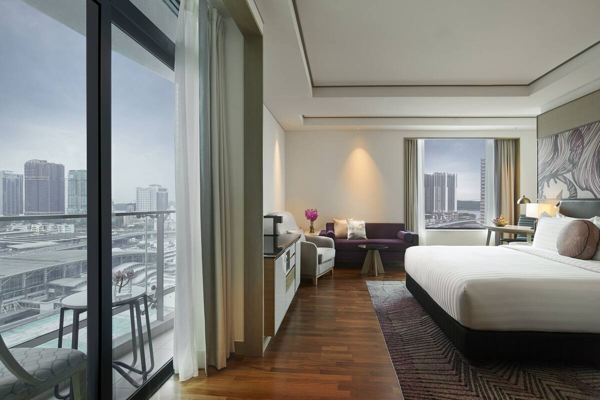 Quarto de hotel moderno com uma cama grande, um sofá, uma mesa pequena e vista para a paisagem urbana pelas grandes janelas.