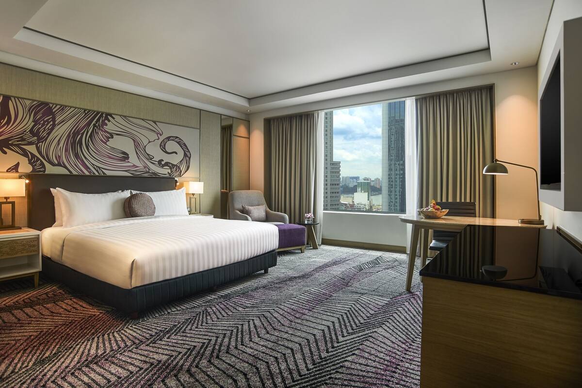 Quarto de hotel moderno com uma cama grande, uma escrivaninha, uma cadeira e vista para a paisagem urbana pelas grandes janelas.