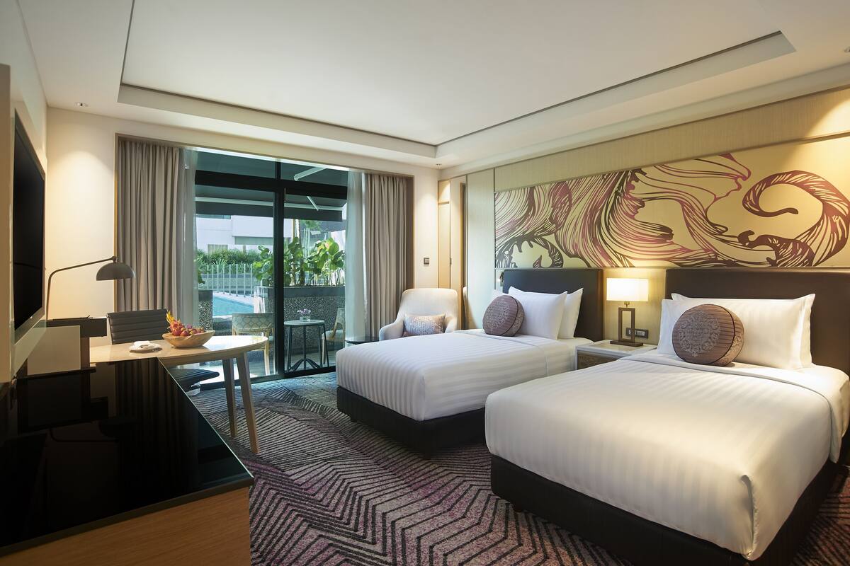 Quarto de hotel com duas camas, uma escrivaninha, uma cadeira e uma grande janela com vista para palmeiras.