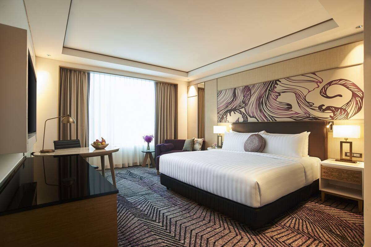Quarto de hotel com uma cama grande, uma escrivaninha com cadeira, televisão e área de refeições com mesa e cadeiras.