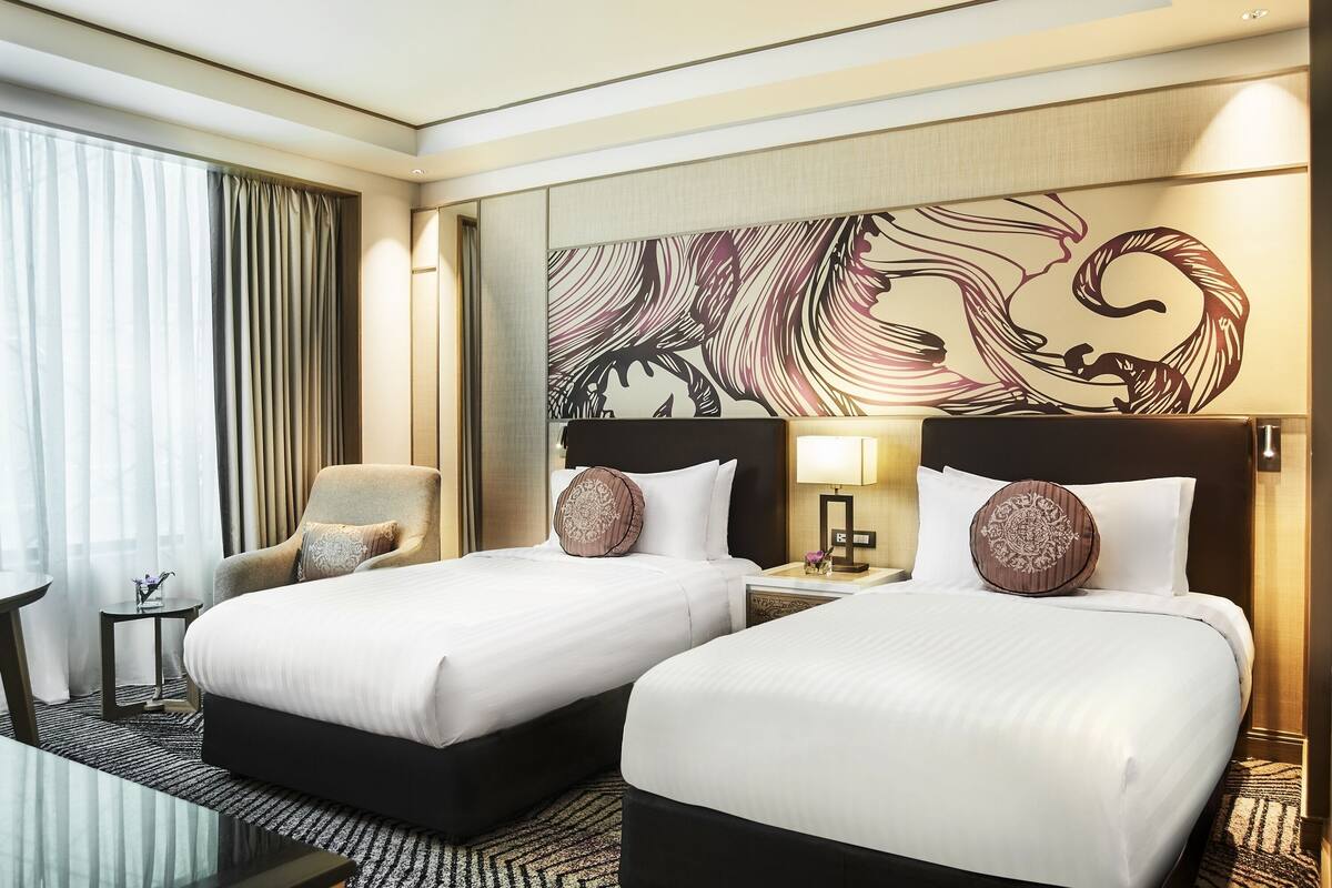 Quarto de hotel com duas camas, uma cadeira, uma mesa pequena e um objeto decorativo na parede.