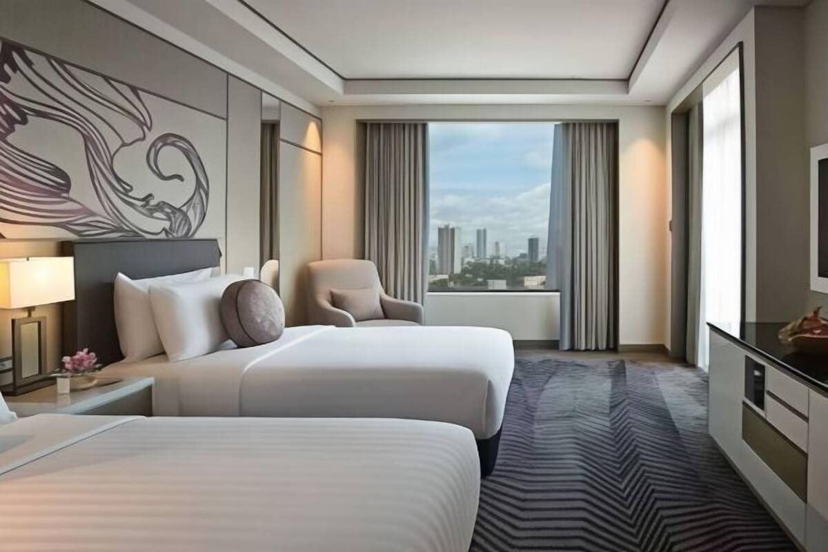 Quarto de hotel com duas camas, uma janela ampla com vista para a cidade, uma obra de arte moderna na parede e uma televisão de tela plana.