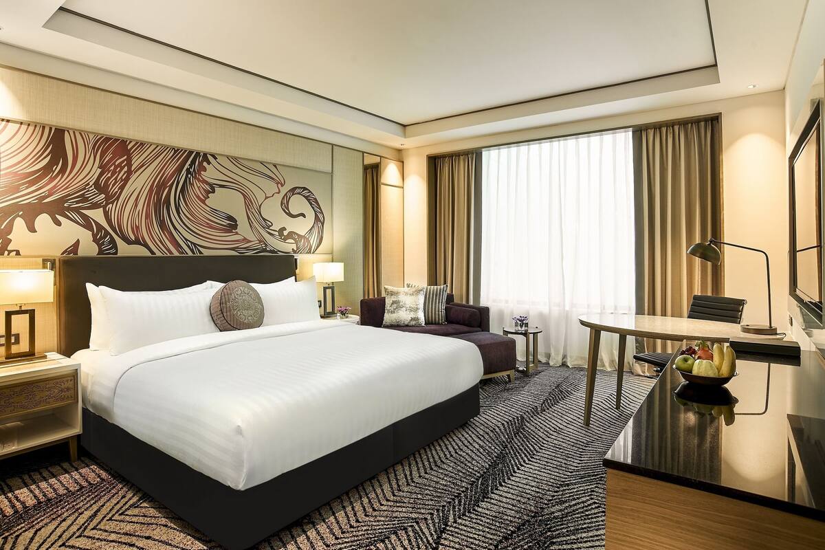 Quarto de hotel com uma cama grande, um sofá, uma mesa de trabalho com abajur e uma televisão.
