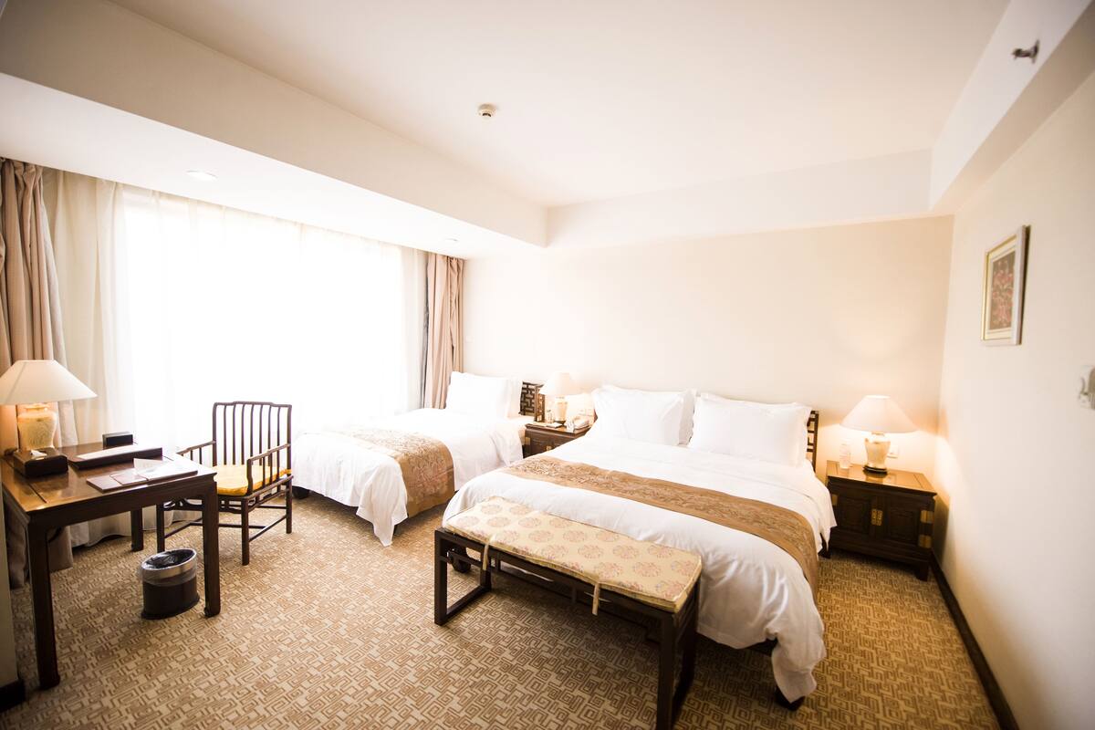 Quarto de hotel com duas camas, uma escrivaninha, uma cadeira, um banco e uma mesa de cabeceira com luminárias.