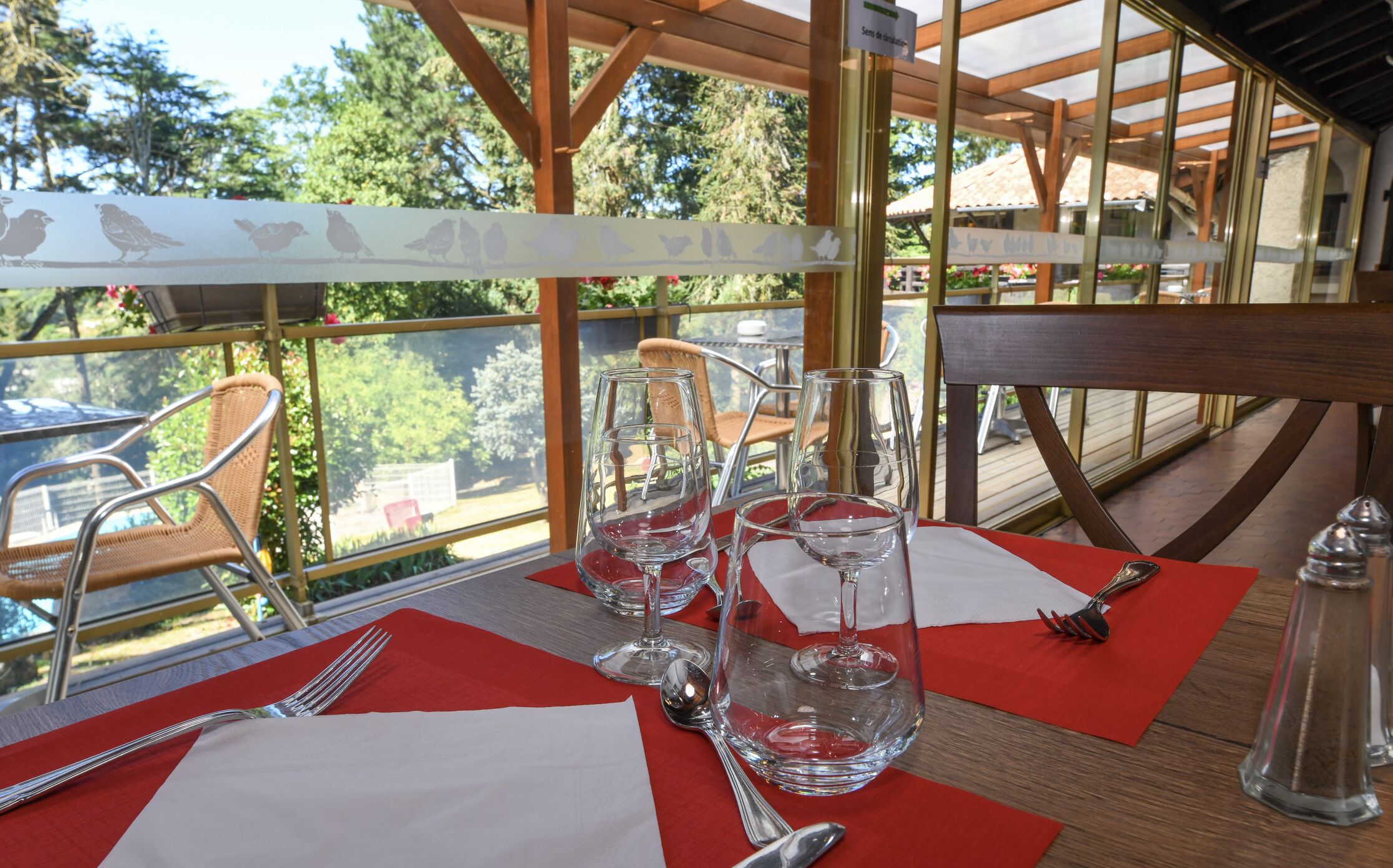 Uma mesa posta com copos de vinho, talheres e guardanapos sobre um tapete vermelho, com vista para um jardim com cadeiras e uma pérgola de madeira.