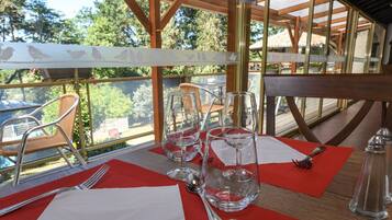 Uma mesa posta com copos de vinho, talheres e guardanapos sobre um tapete vermelho, com vista para um jardim com cadeiras e uma pérgola de madeira.