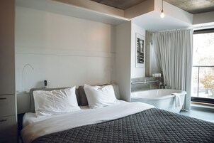 Quarto de hotel moderno com cama grande, banheira integrada e uma ampla janela com cortinas.