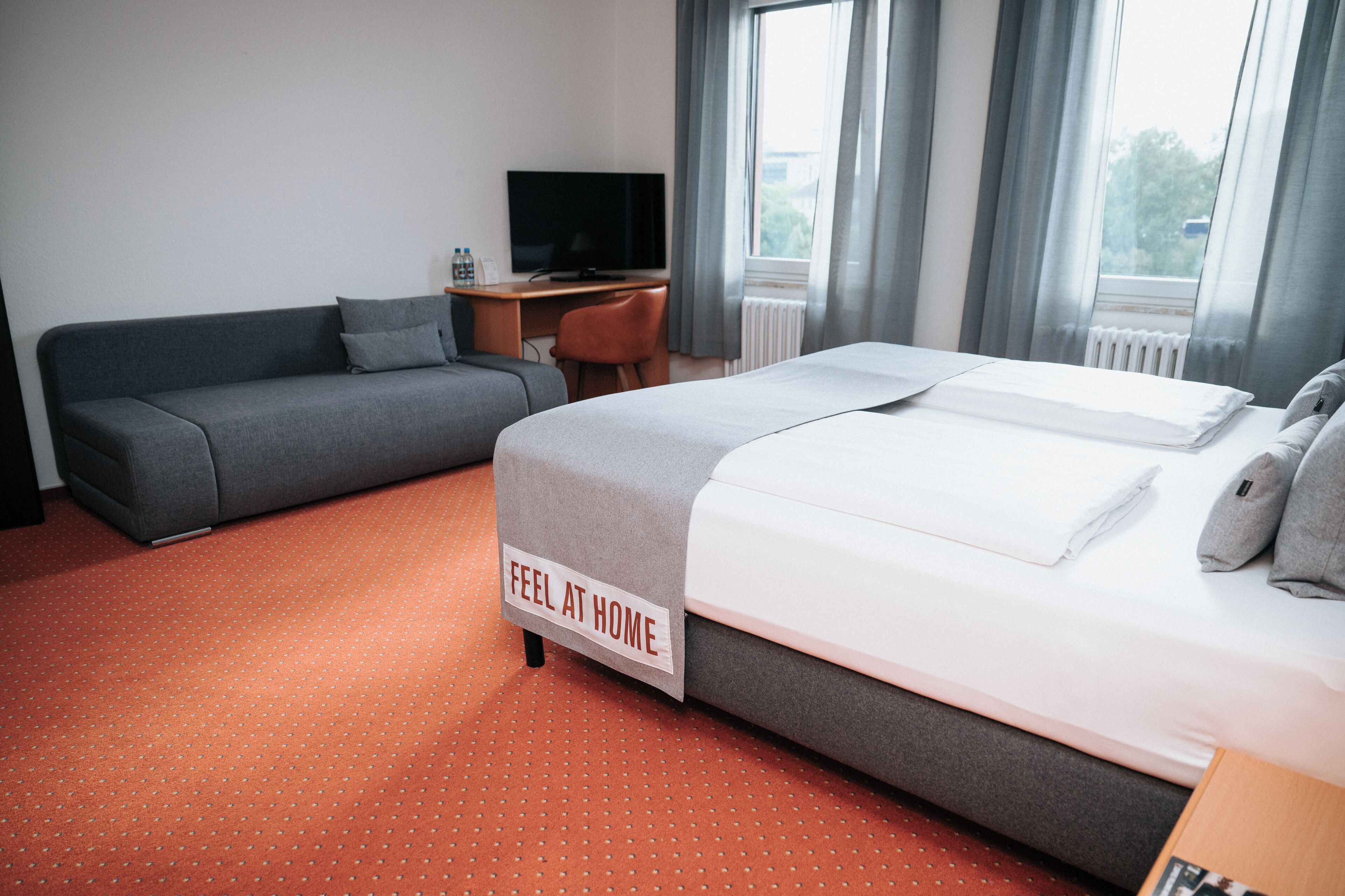 Quarto de hotel com cama, sofá, escrivaninha com televisão e janela com cortinas.
