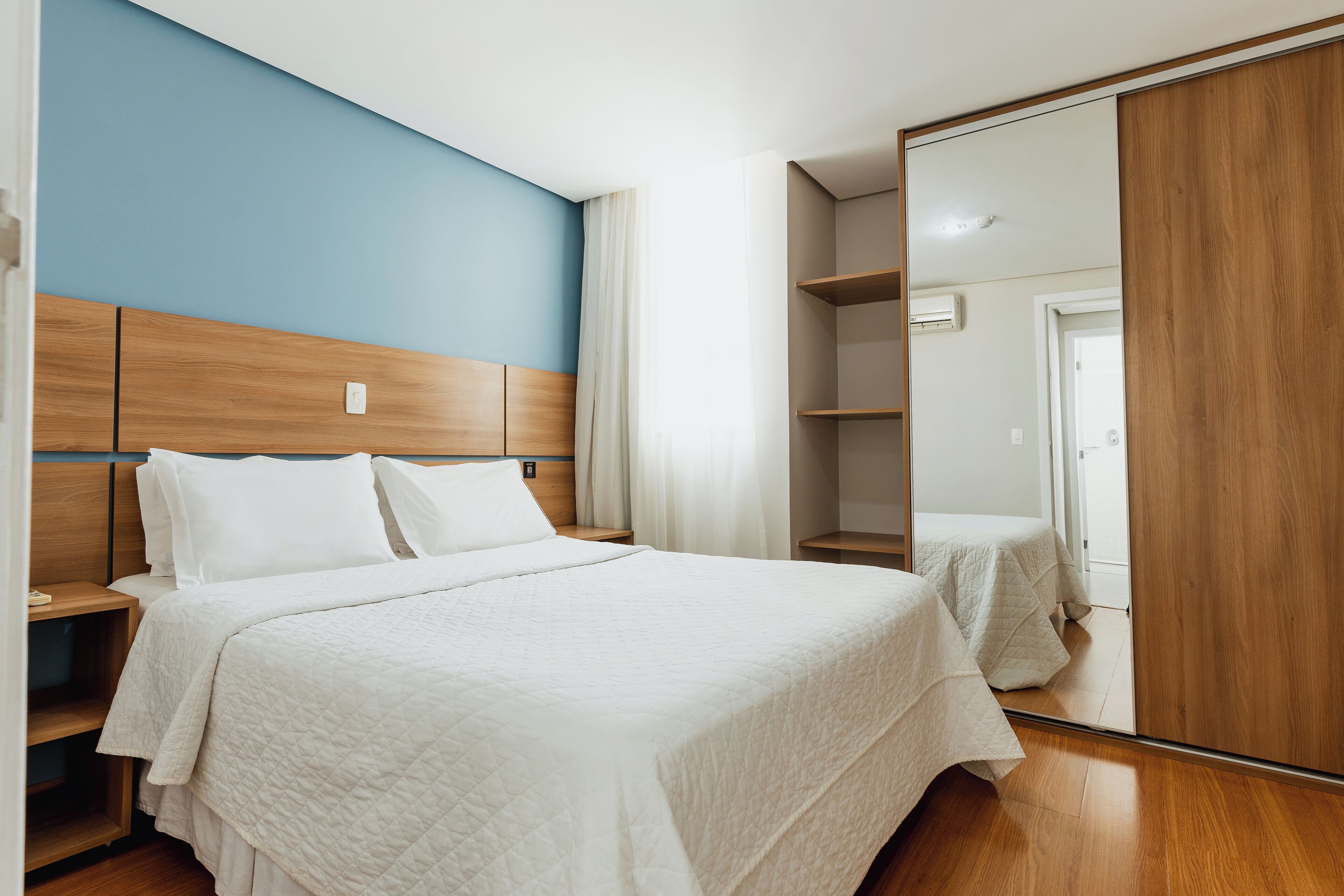 Quarto de hotel com uma cama grande, cabeceira de madeira, um guarda-roupa e um espelho.