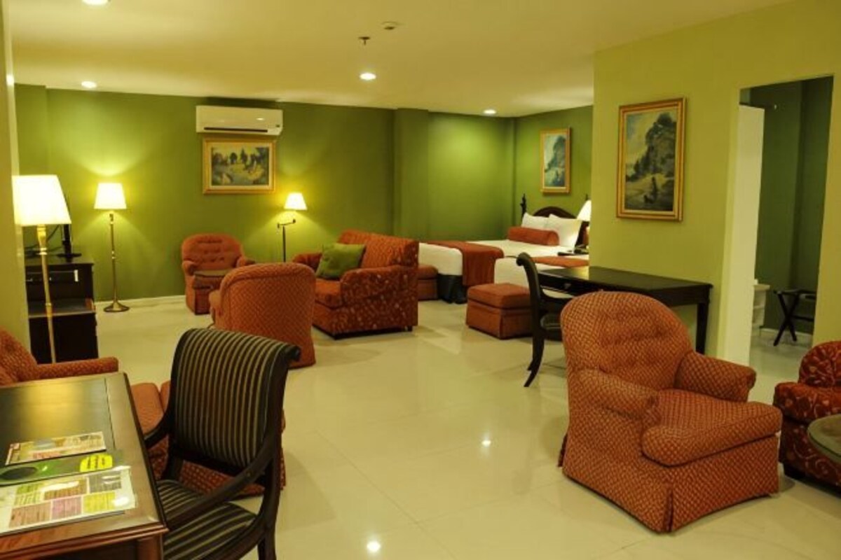 Quarto de hotel com paredes verdes, um sofá, uma mesa de jantar e duas luminárias.