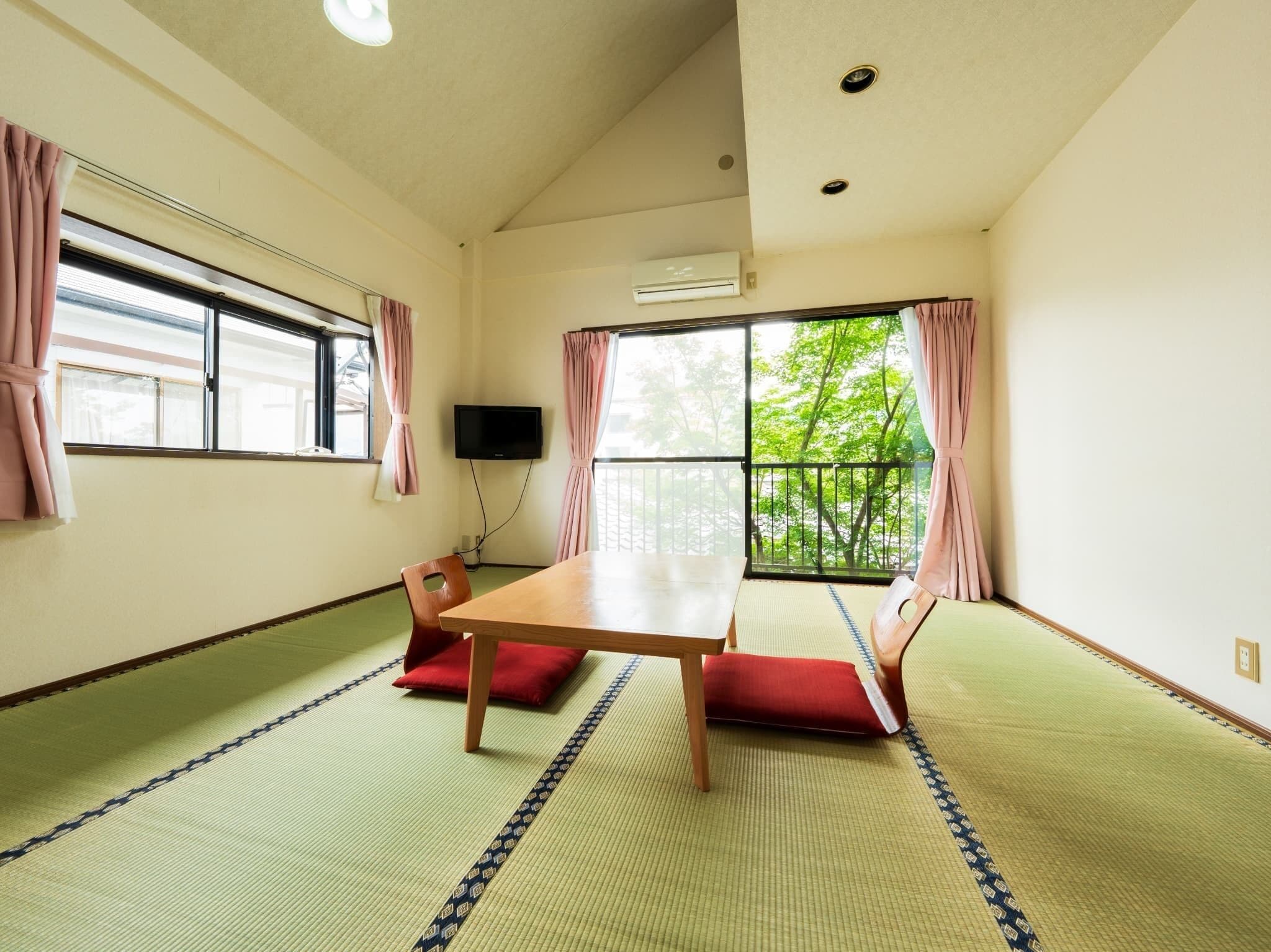Quarto com esteiras de tatame, uma mesa de madeira, cadeiras, uma TV e janelas com cortinas.