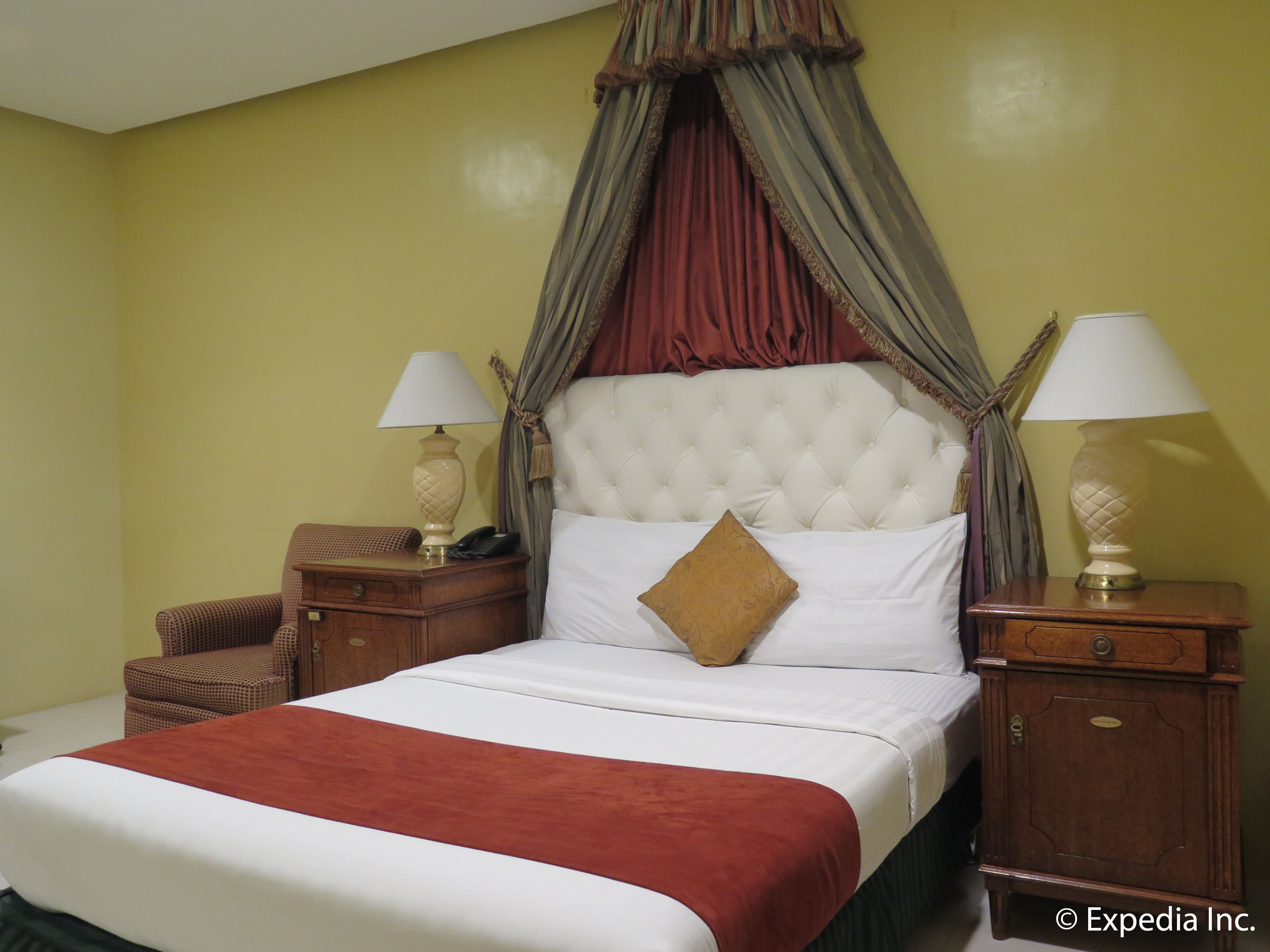 Uma cama com dossel e cabeceira estofada, duas mesas de cabeceira com luminárias, um criado-mudo de madeira e um cobertor vermelho.