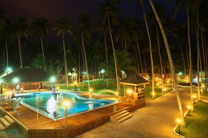Área de piscina à noite com deck de madeira, palmeiras e estruturas iluminadas.