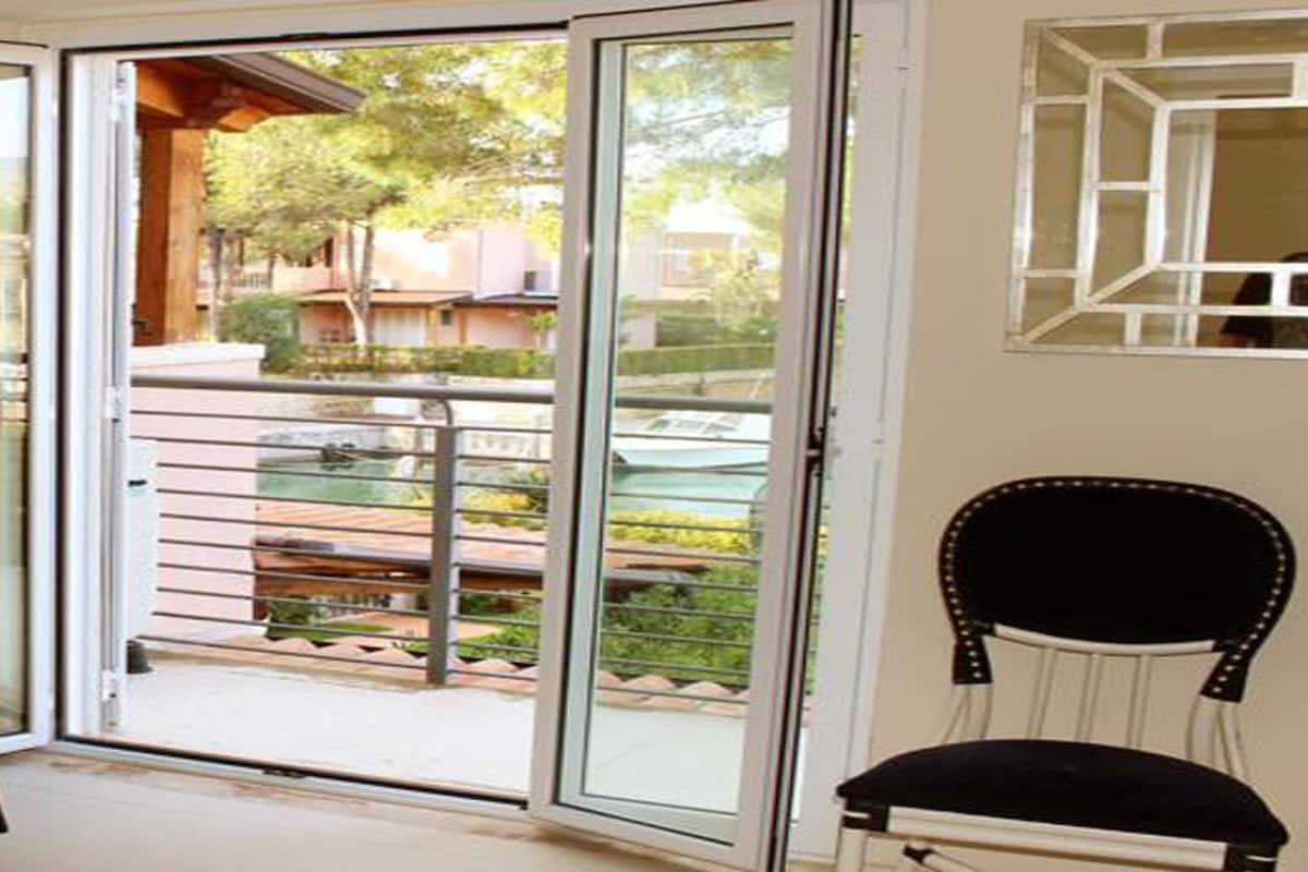 Quarto com cama, uma cadeira, uma mesinha e uma porta de vidro deslizante para uma varanda com vista para um jardim e edifícios.