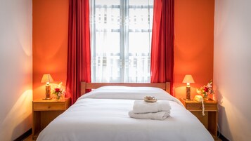 Quarto de hotel com uma cama grande, cortinas vermelhas, duas mesas de cabeceira com abajures e uma janela com vista para edifícios.