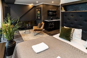 Quarto moderno com uma cama grande, área de estar com uma poltrona, pequena cozinha e uma planta em vaso.