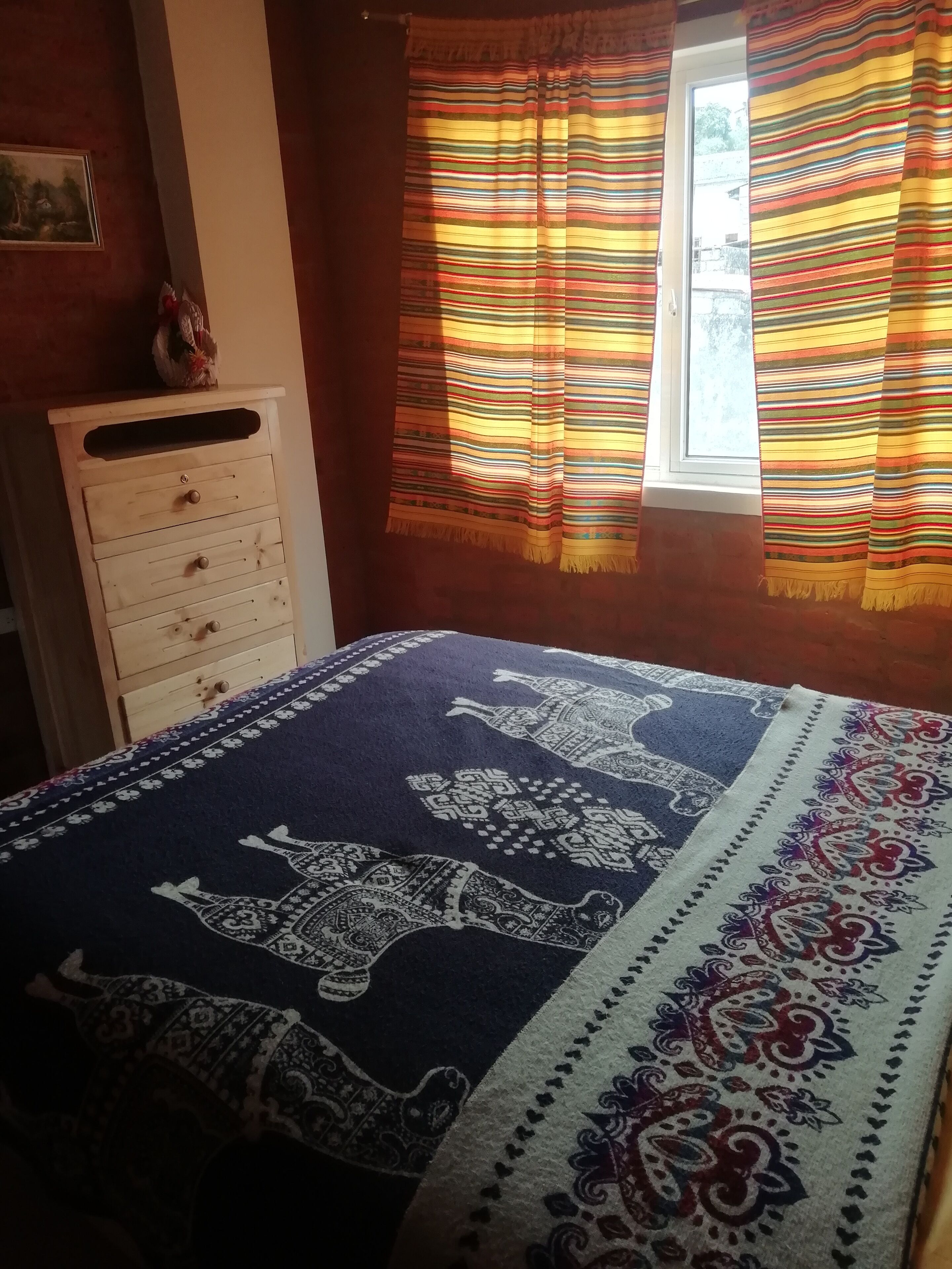 Um quarto com uma cama coberta por um cobertor estampado, uma cômoda de madeira com gavetas e uma janela com cortinas listradas.