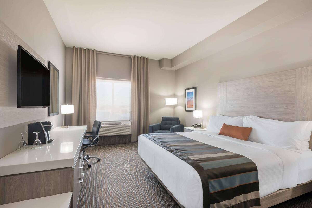 Quarto de hotel com cama, escrivaninha com cadeira, televisão, janela com cortinas e luminária na parede.
