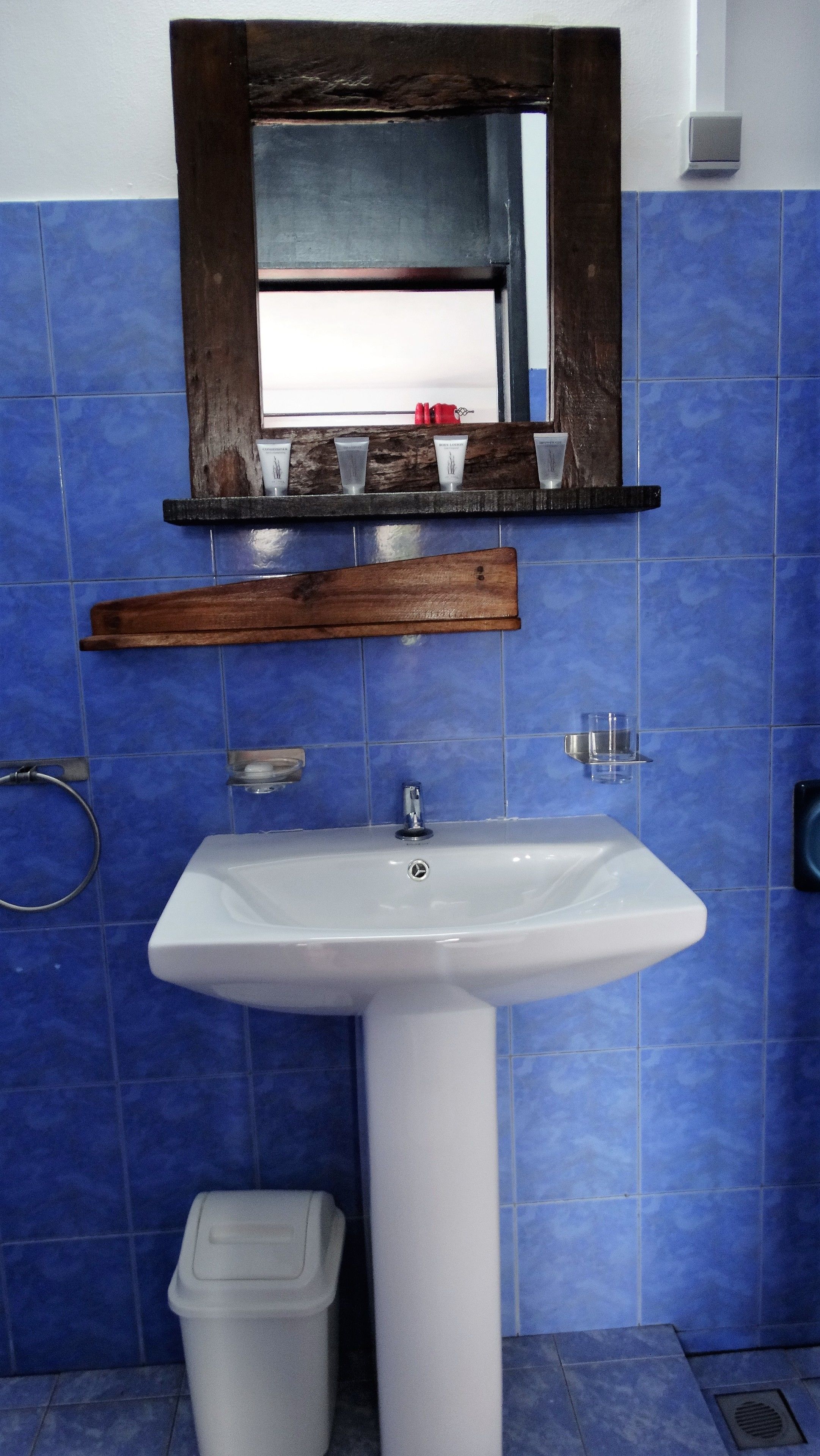 Casa de banho com lavatório branco, azulejos azuis, prateleira de madeira com artigos de higiene e um cesto de lixo.