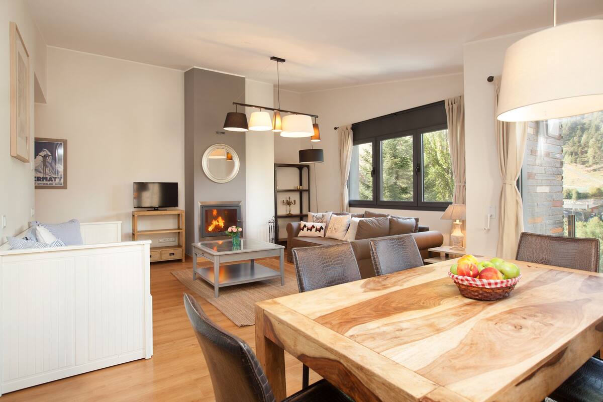 Sala de estar moderna com lareira, mesa de jantar com cesta de frutas e uma janela grande com vista para o exterior.
