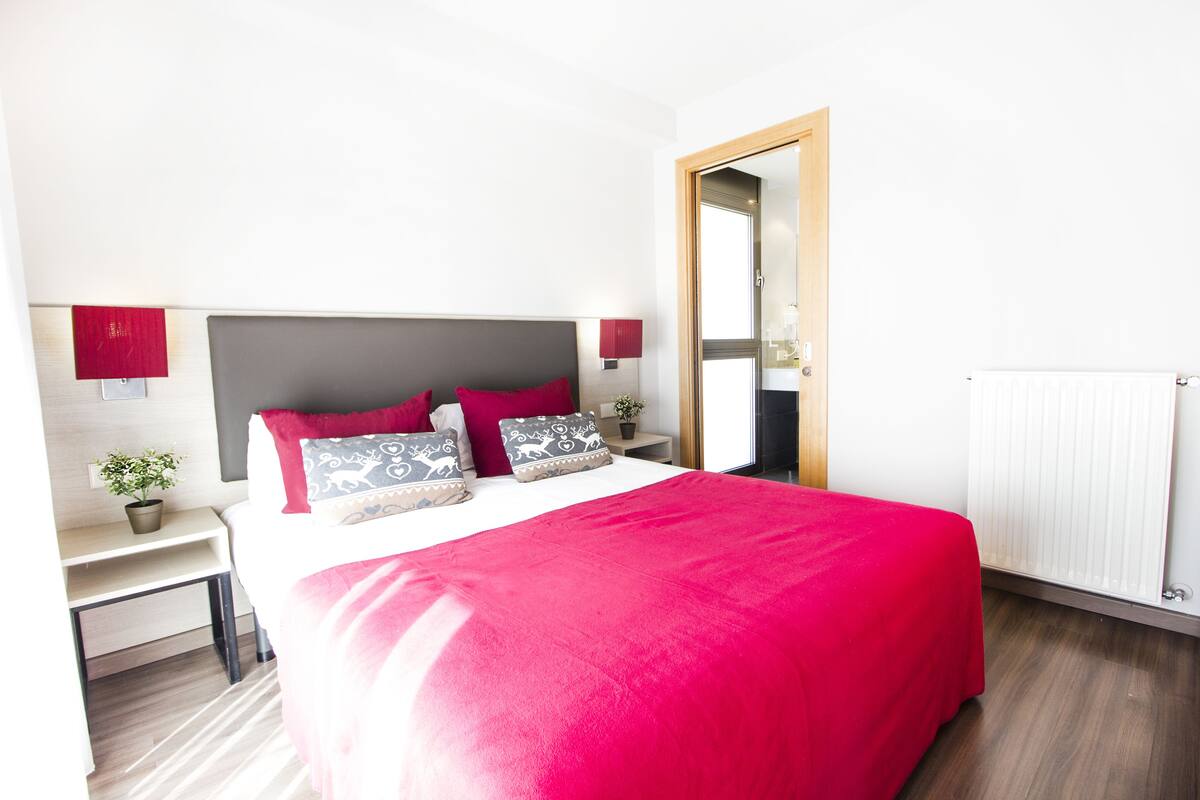 Quarto de hotel moderno com uma cama grande, uma mesa de cabeceira, uma planta em vaso e uma janela com vista.