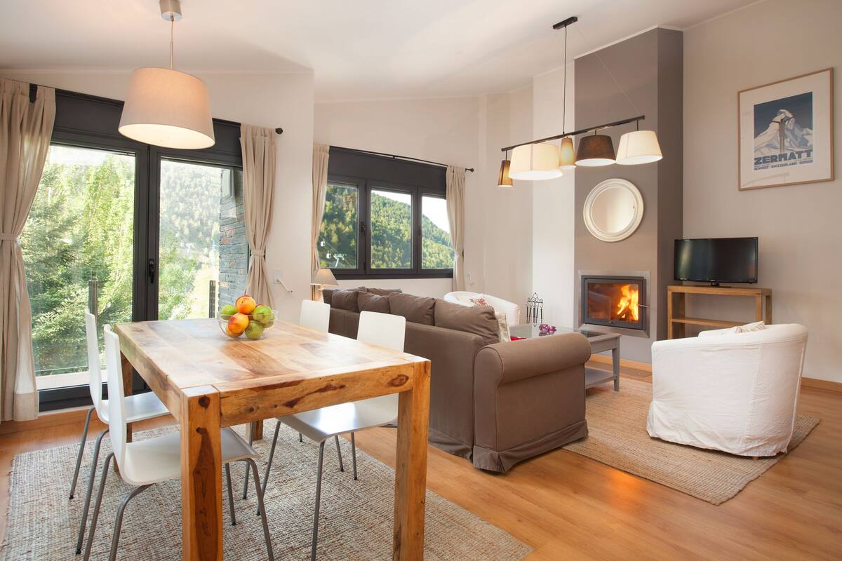 Uma sala de estar moderna com uma mesa de jantar de madeira, cadeiras brancas, um sofá marrom, uma poltrona branca e uma lareira.