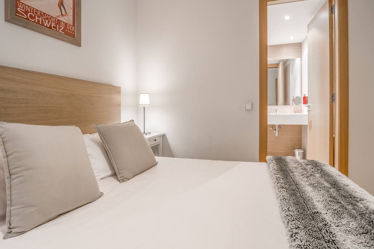 Quarto de hotel com cabeceira de madeira, cama com duas almofadas, mesa de cabeceira com abajur e banheiro visível através de uma porta aberta.