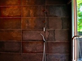 Casa de banho com azulejos castanhos escuros, chuveiro com barra de metal cromado e janela com vista para vegetação.