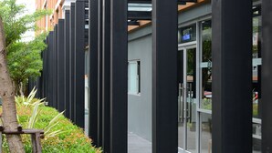 Um edifício moderno com entrada de vidro, colunas verticais pretas e um jardim bem cuidado com um banco de madeira.